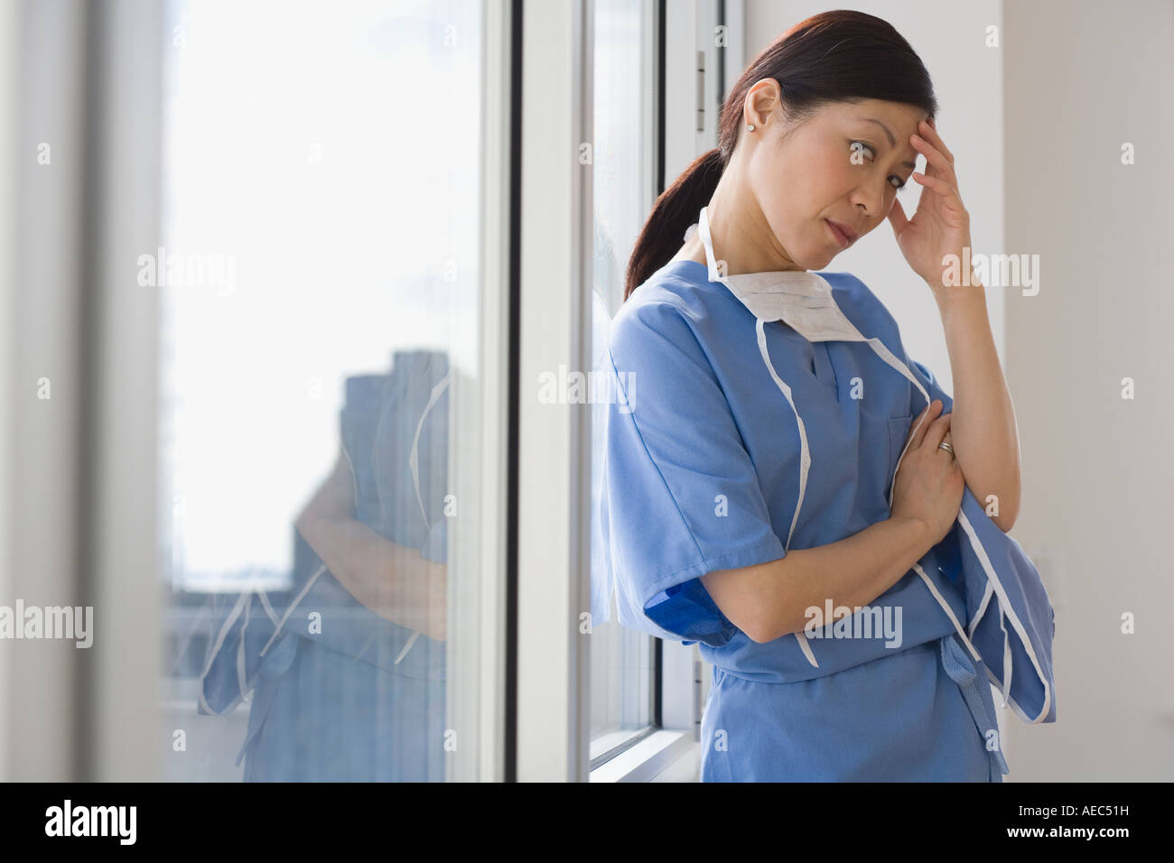 Asian female doctor leaning against window with hand on forehead Stock ...