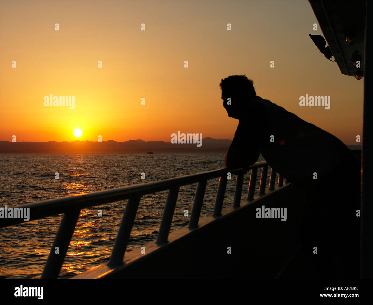 Man silhouetted by sunset, leaning on safety rail on deck of a ship at ...