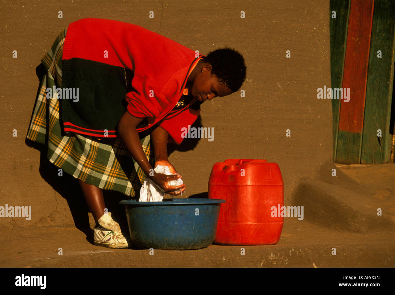 A woman washing clothing in a plastic bowl outside a mud-walled ...