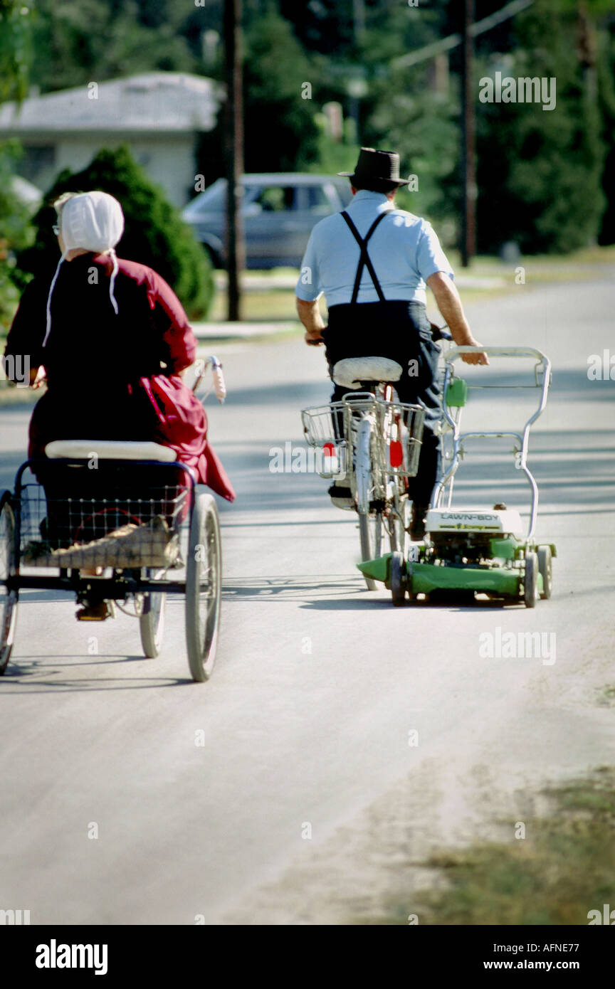 Amish Male and female ride down the street on bicycles dragging a lawn ...