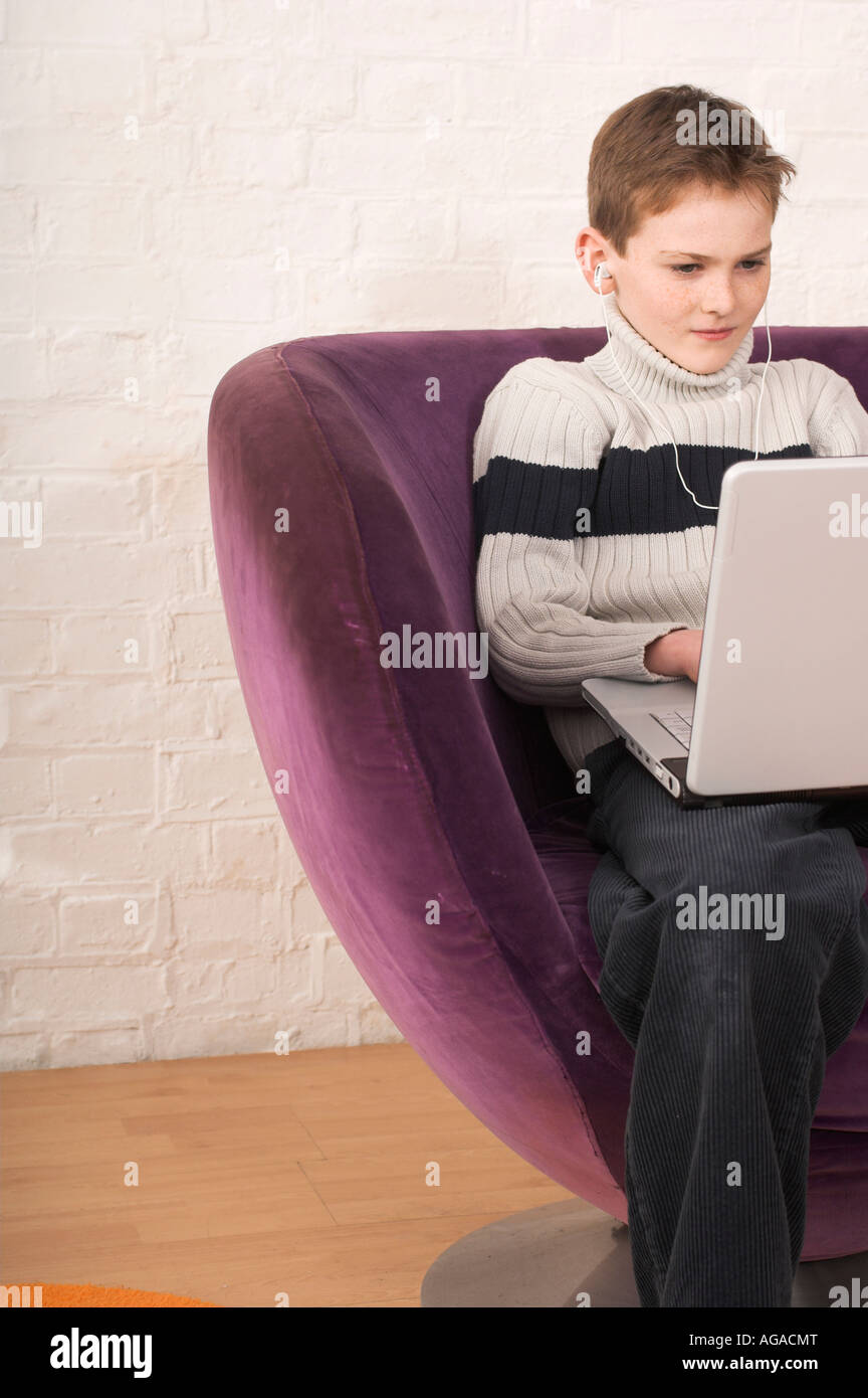 portrait of young boy sitting in arm chair with earphones and laptop ...