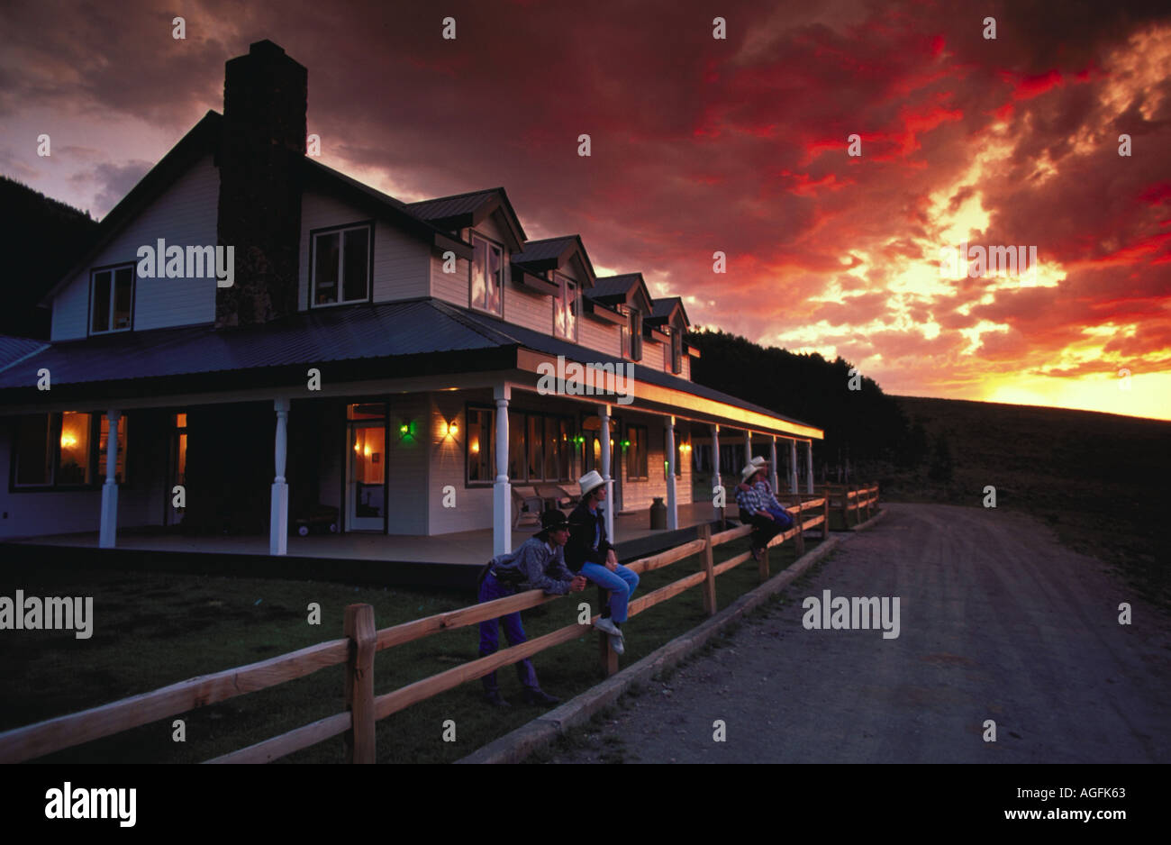 Three people sitting and one standing along a fence in front of a ranch ...