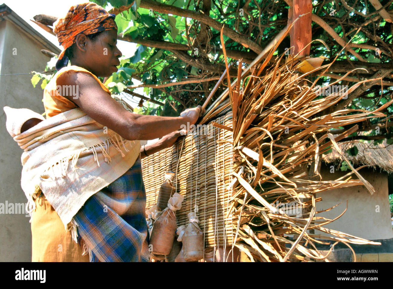 An African lady with a baby on her back weaves a reed mat the ...