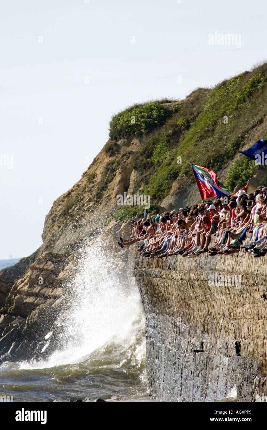 Spectators watching traineras regatta as sea crashes on sea wall below ...