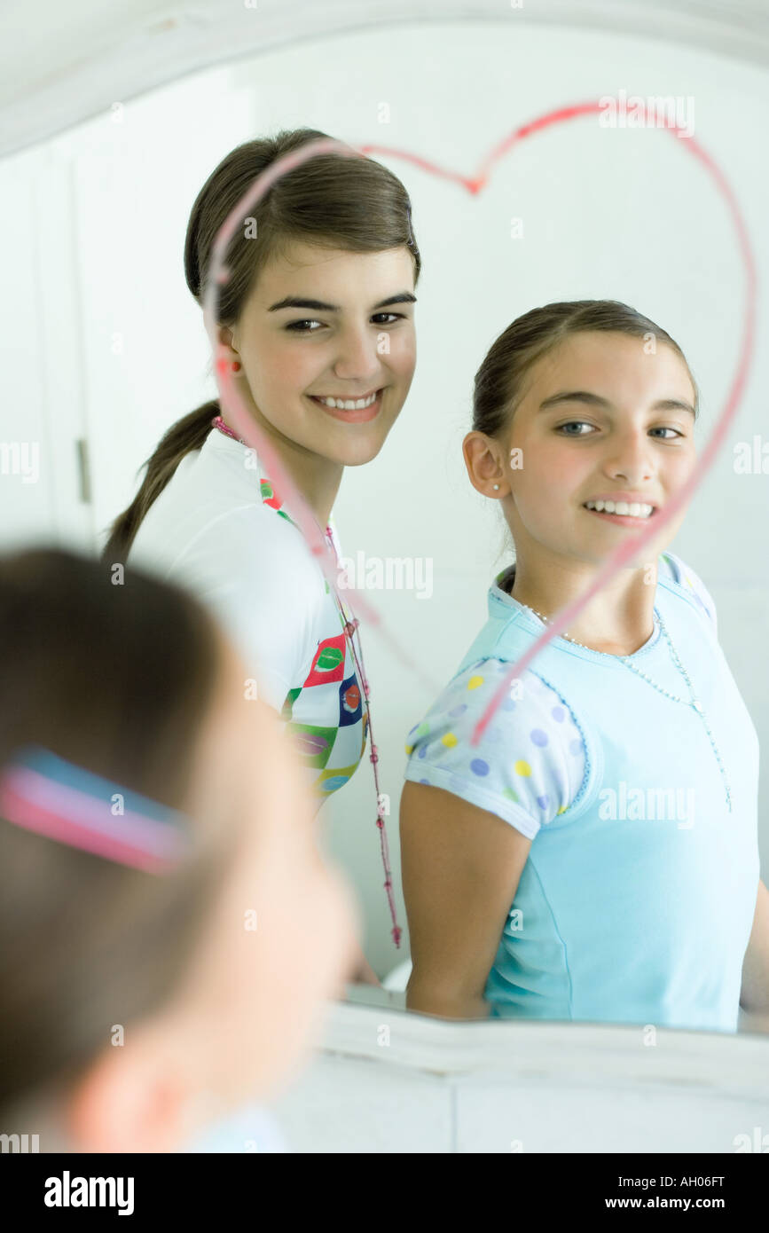 Two young female friends looking in mirror, heart drawn on mirror with ...