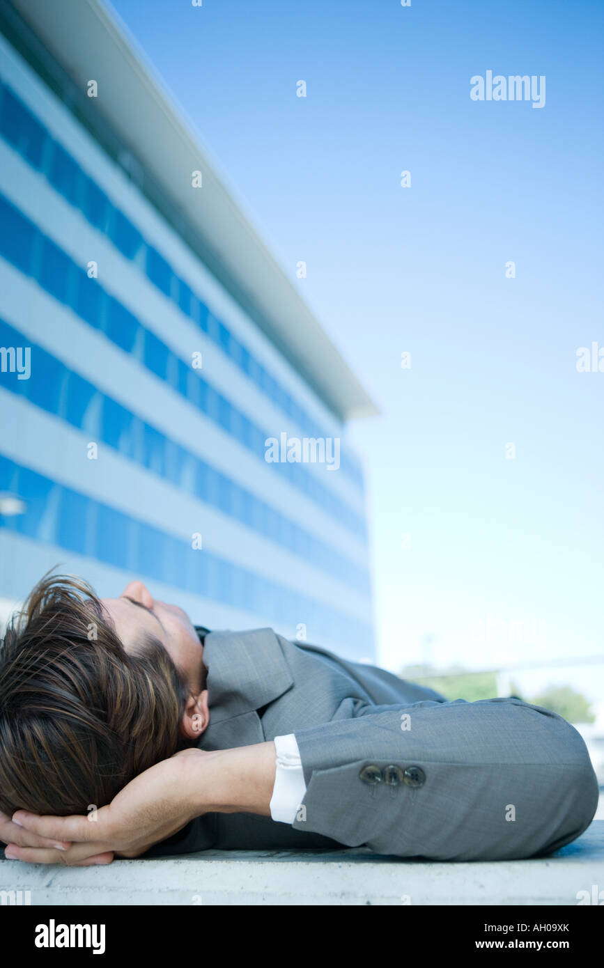 Businessman lying on ground outdoors, hands behind head, cropped view ...