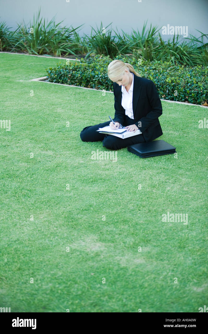 Young businesswoman sitting on the ground outdoors, writing on ...