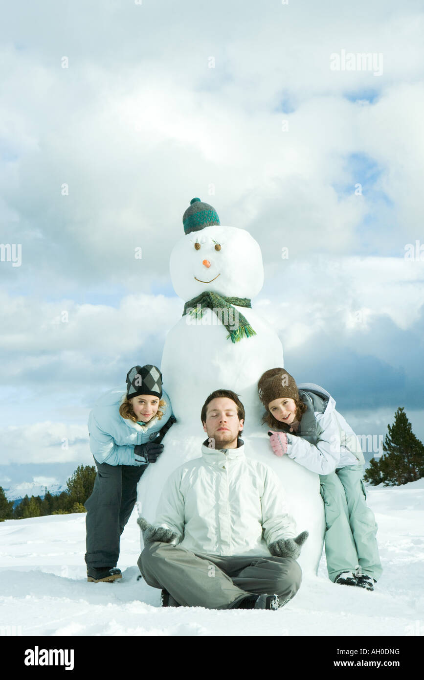 Young man meditating in front of snowman, two sisters crouching behind ...
