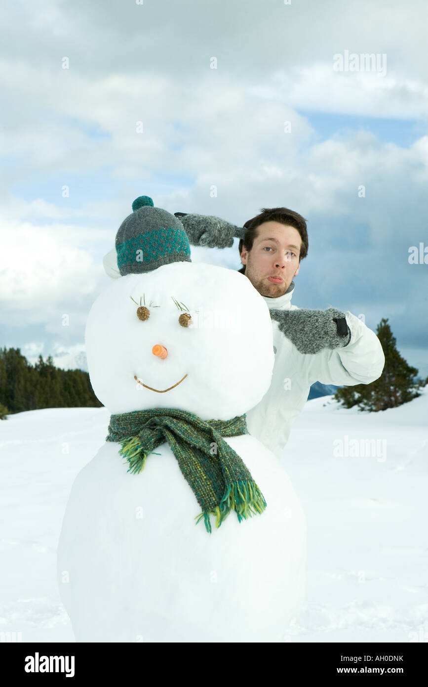 Young man standing behind snowman, pointing, making faces at camera ...