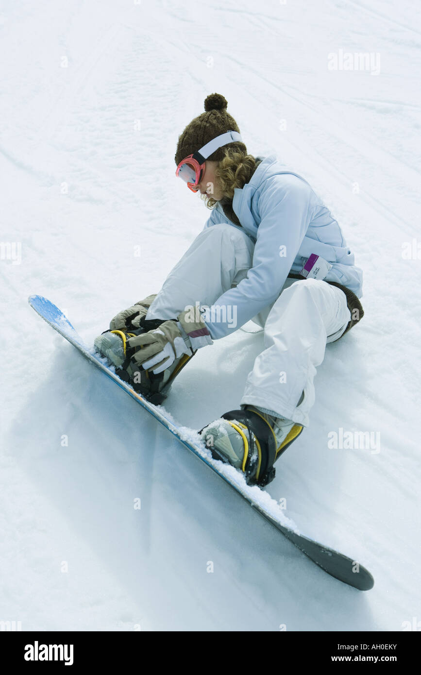 Teenage girl sitting on the ground, putting on snowboard, full length ...
