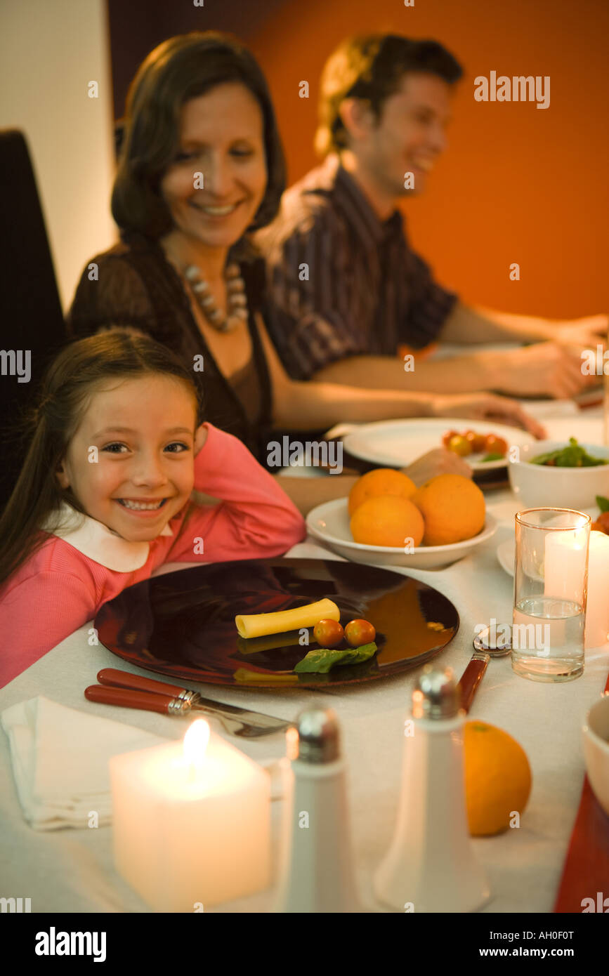 Girl sitting at dinner table, smiling at camera, adults in background ...