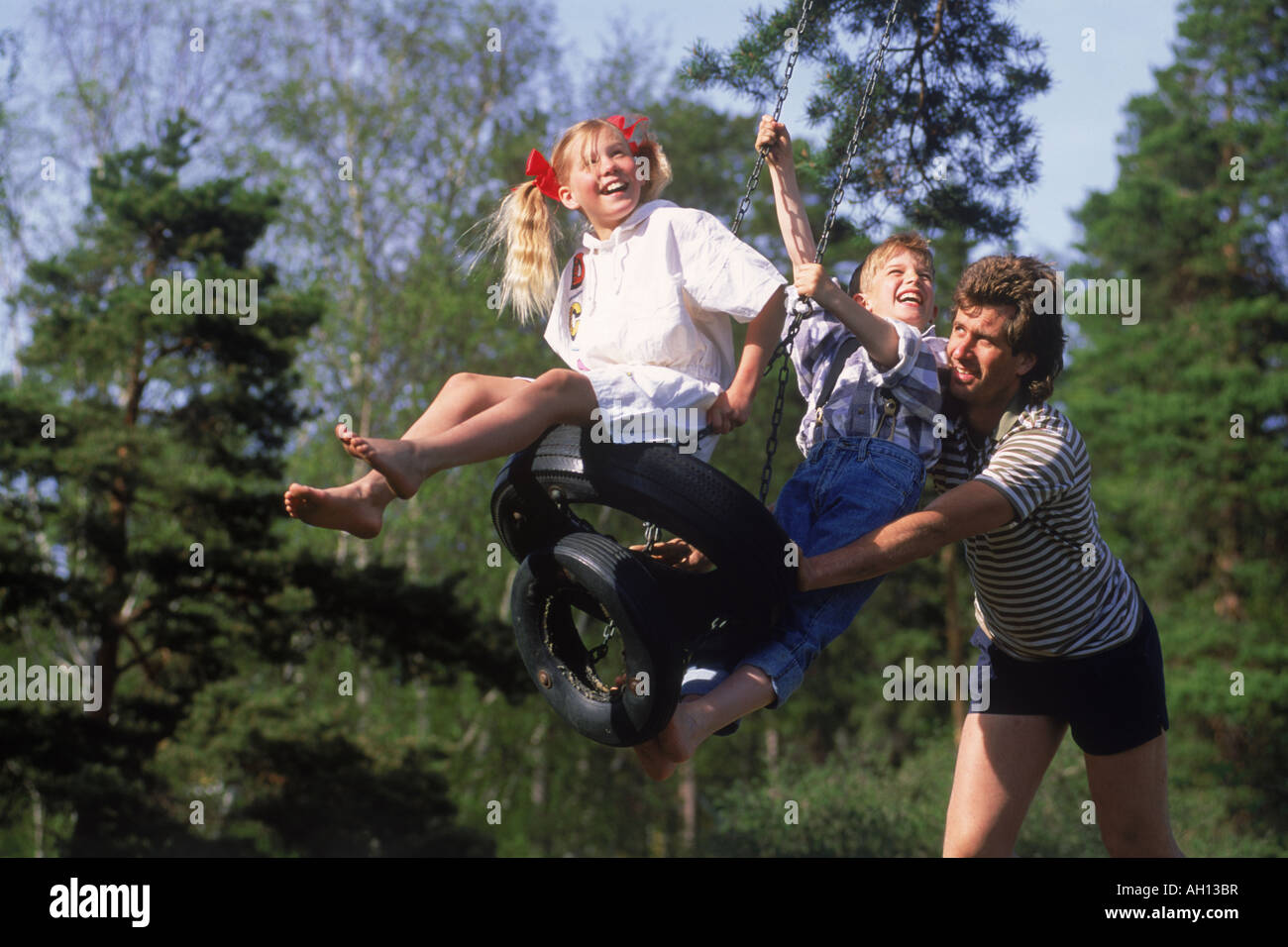 Father helping push swing holding son and daughter on summer day in ...