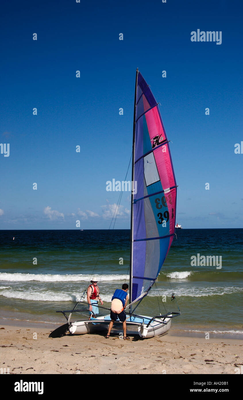 Two Caucasian men launch a blue and pink sailboat into the sparkling ...