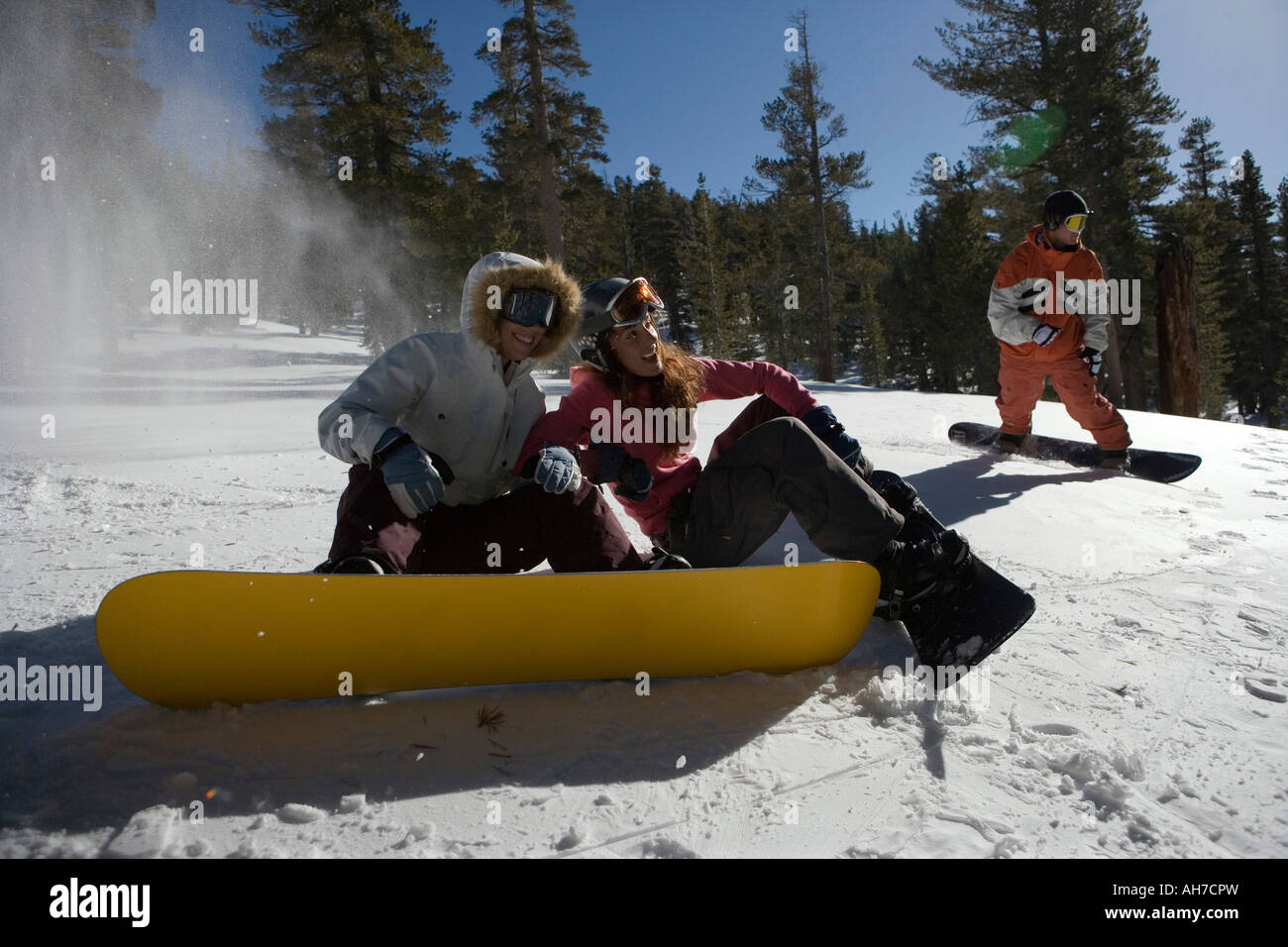 Two young women sitting on snow with a young man snowboarding beside ...