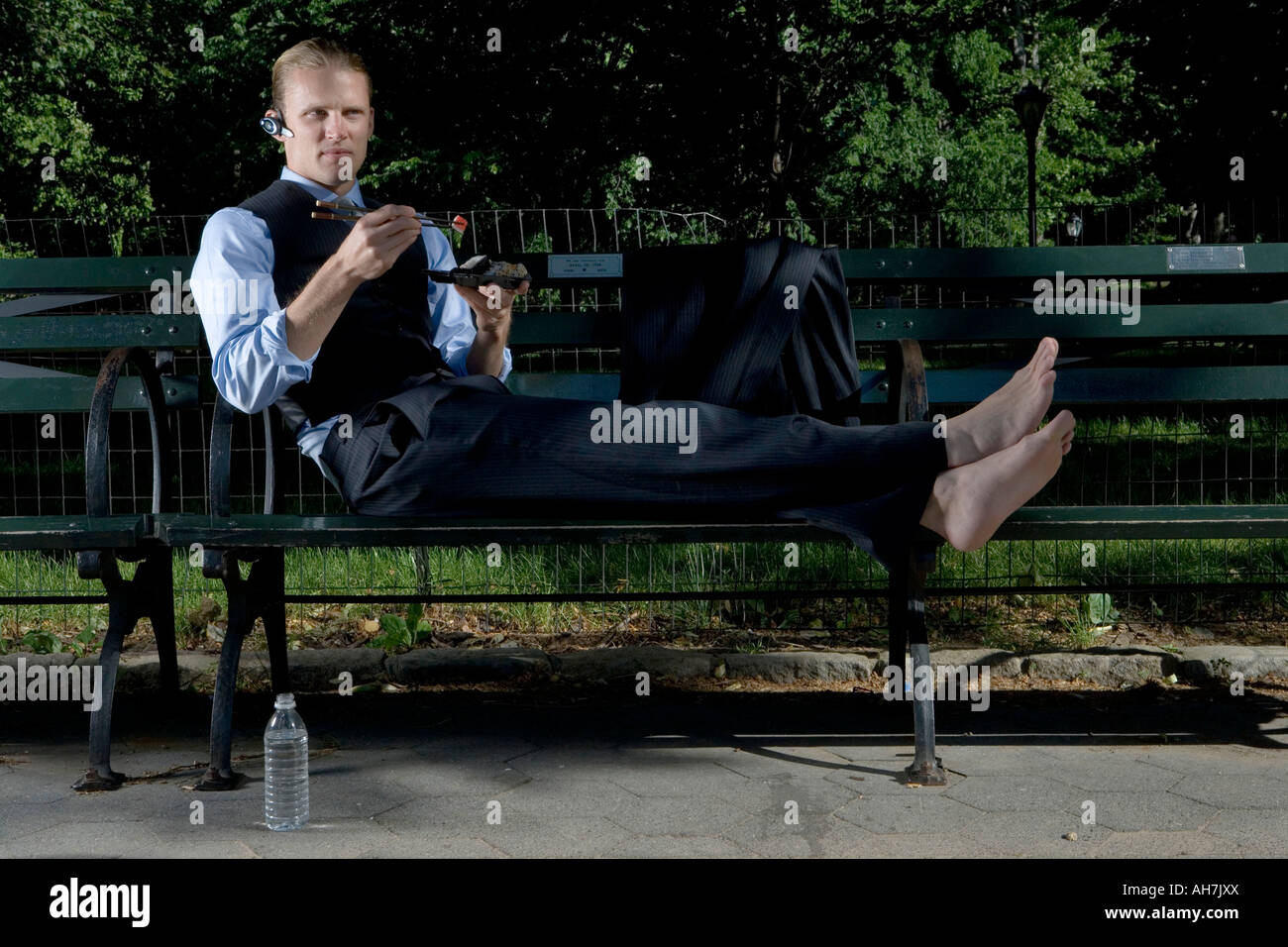 Businessman reclining on a park bench and eating food with chopsticks ...