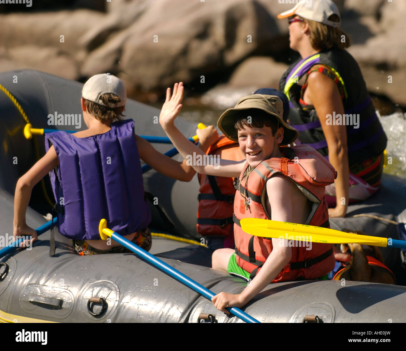 A young boy paddling a whitewater raft with his family smiles and waves ...