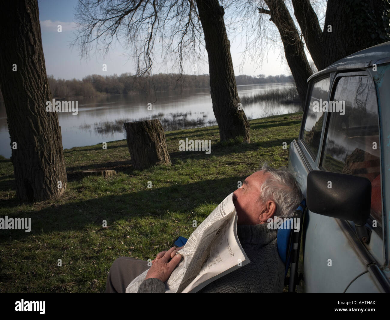 Senior man resting by car on riverside, holding newspaper, eyes closed ...