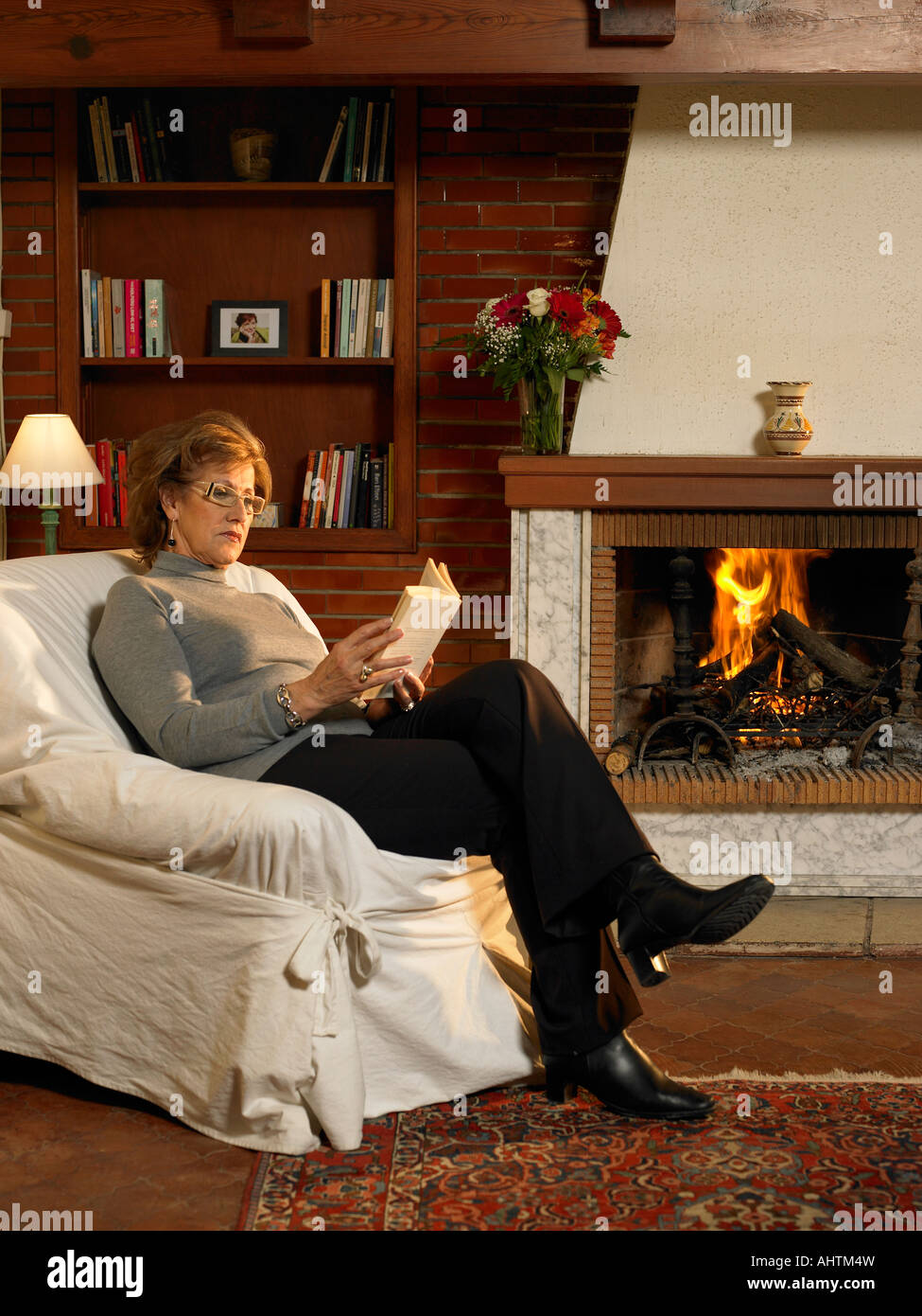 Senior woman reading book by fire in living room Stock Photo - Alamy