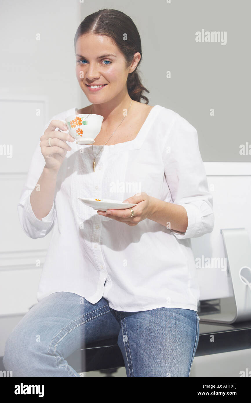 Smiling woman sitting on desk drinking tea Stock Photo - Alamy