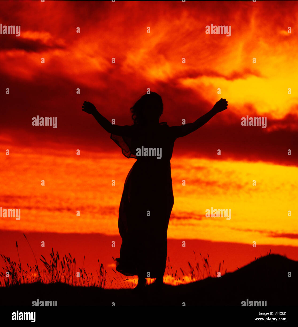 Sunset silhouette of woman in long dress gazing at sunset with arms ...