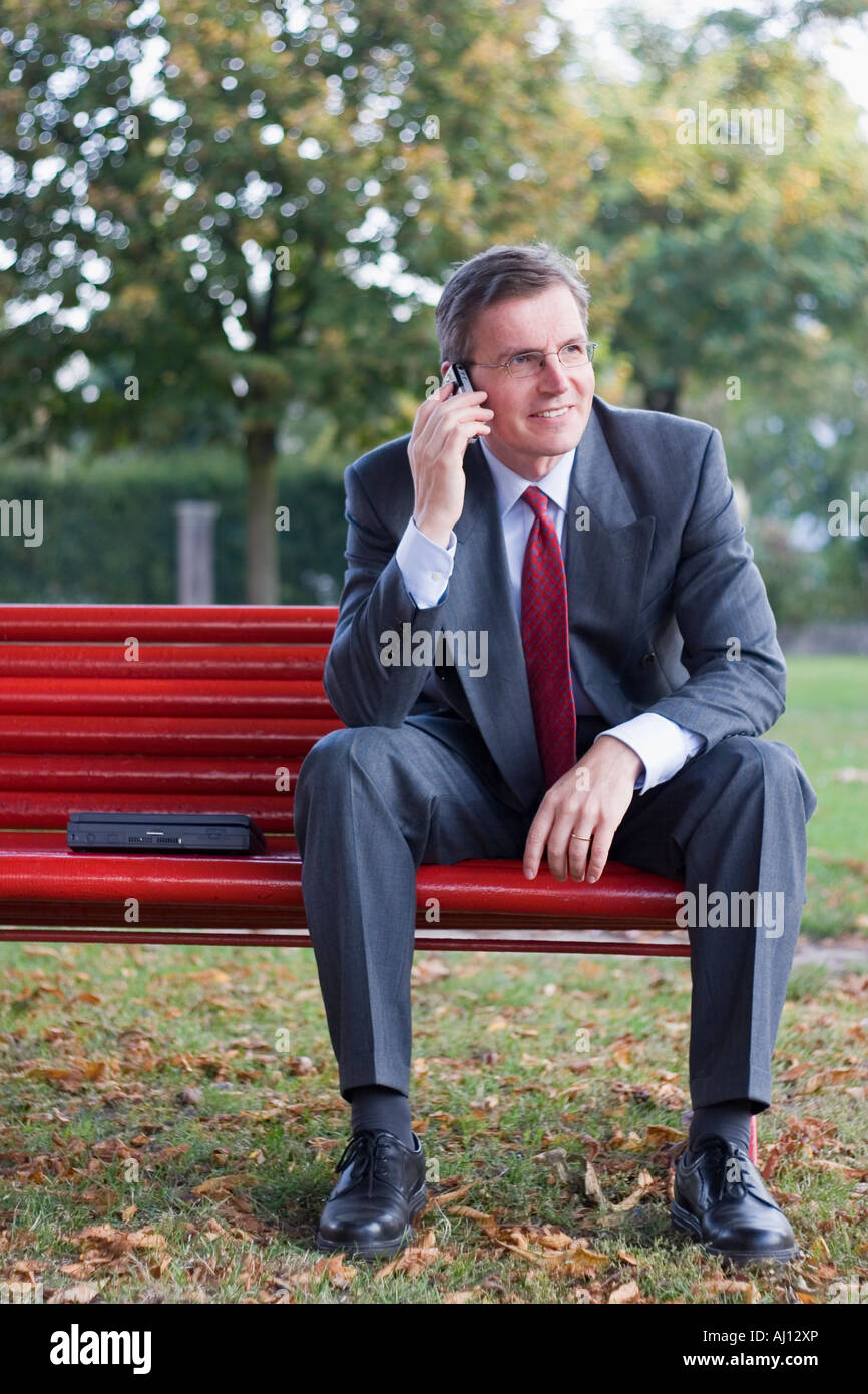 Friendly businessman with cell phone sitting on a red bench in a park ...