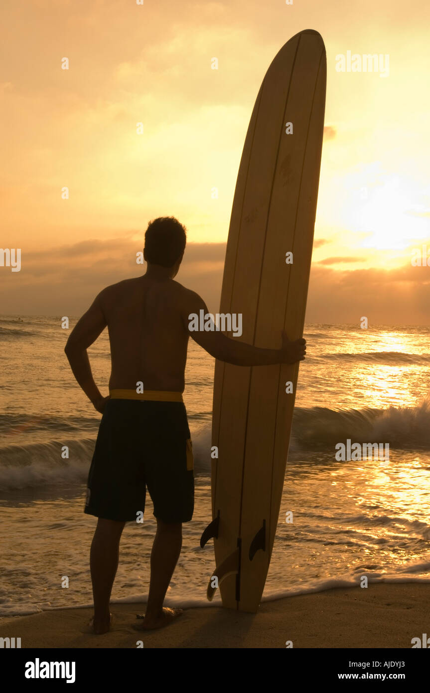 Surfer standing on beach, holding surfboard, watching sunset, back view ...