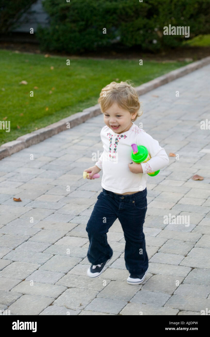Young girl running along a driveway with water bottle and toy doll in ...