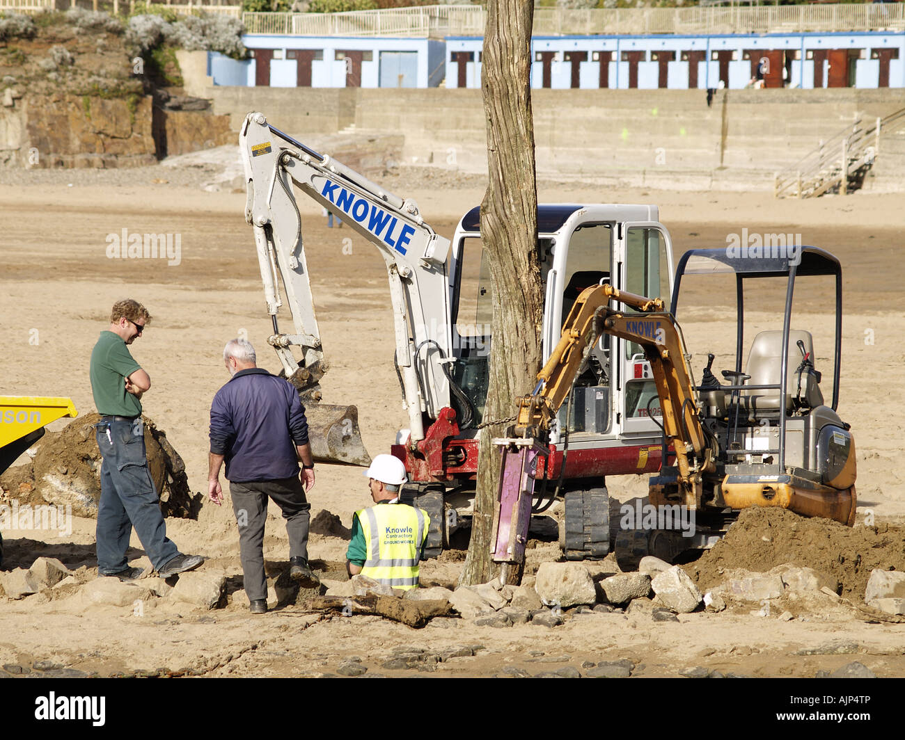 Three workmen using plant machines to dig out a post on a sandy beach ...