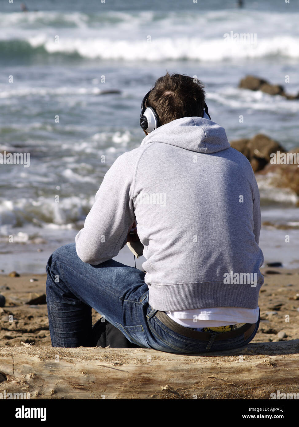 Back of a teenage boy sitting on a log on the beach with headphones on ...
