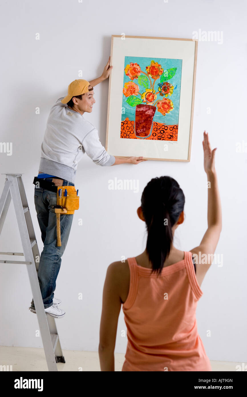Rear view of a woman helping a young man adjust a painting on a wall ...