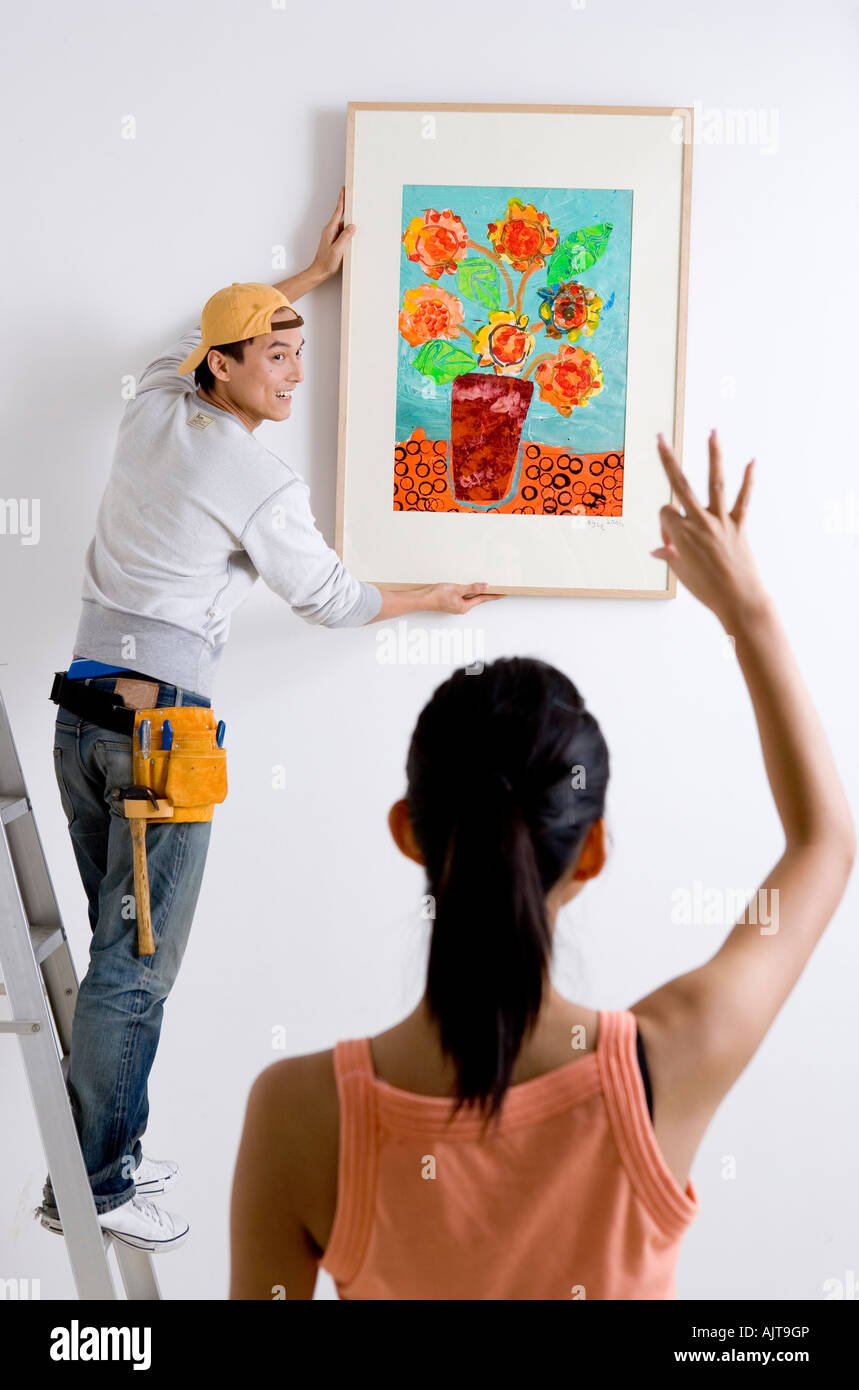 Rear view of a woman helping a young man adjust a painting on a wall ...