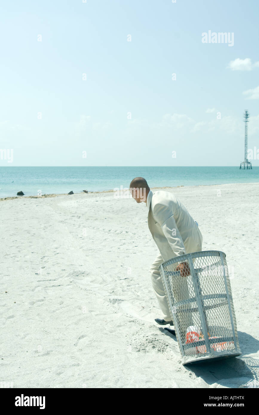 On beach, man in suit pulling garbage can towards ocean, side view ...
