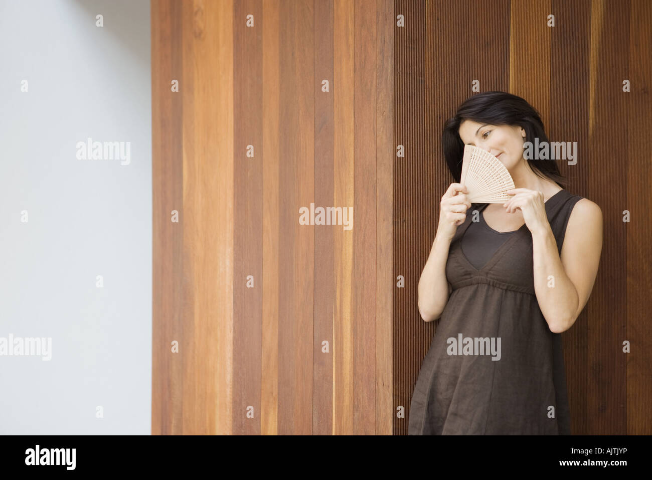 Brunette woman holding up fan in front of face, smiling, looking away ...