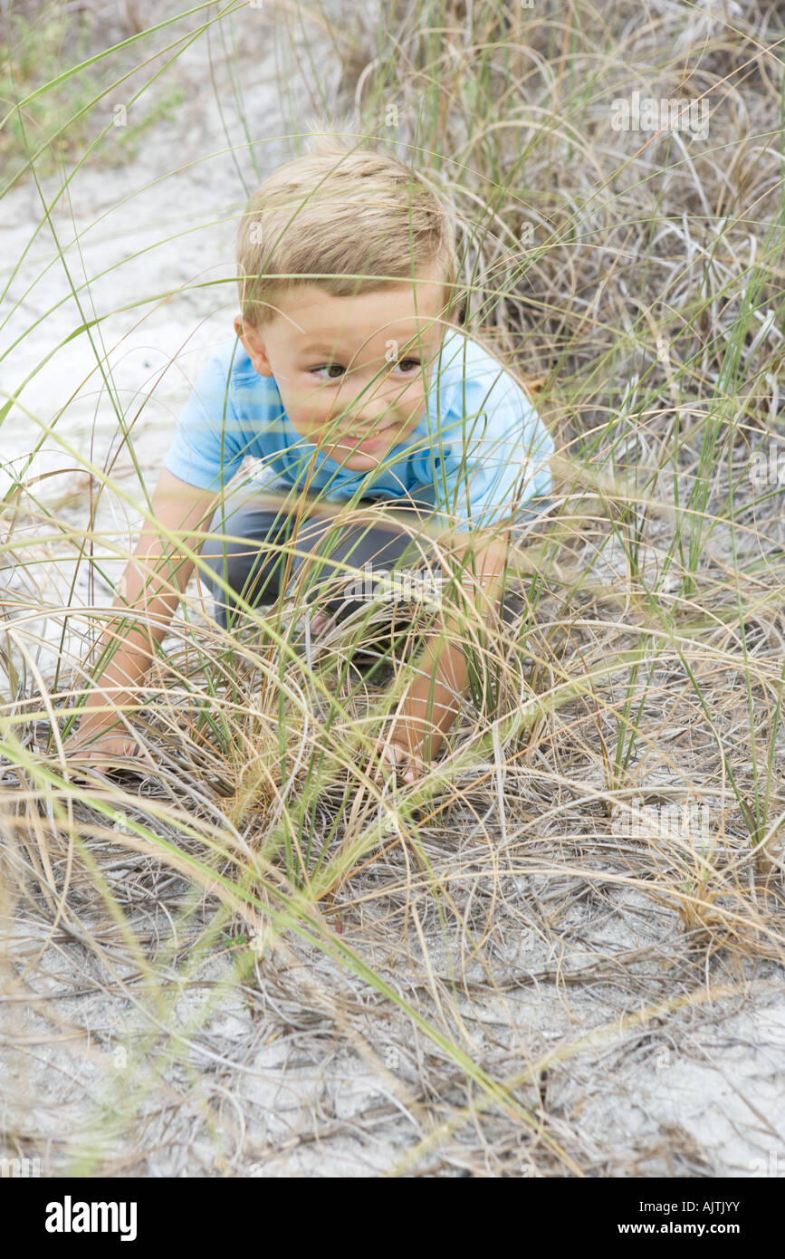 Boy crouching in dune grass, looking away mischievously, full length ...