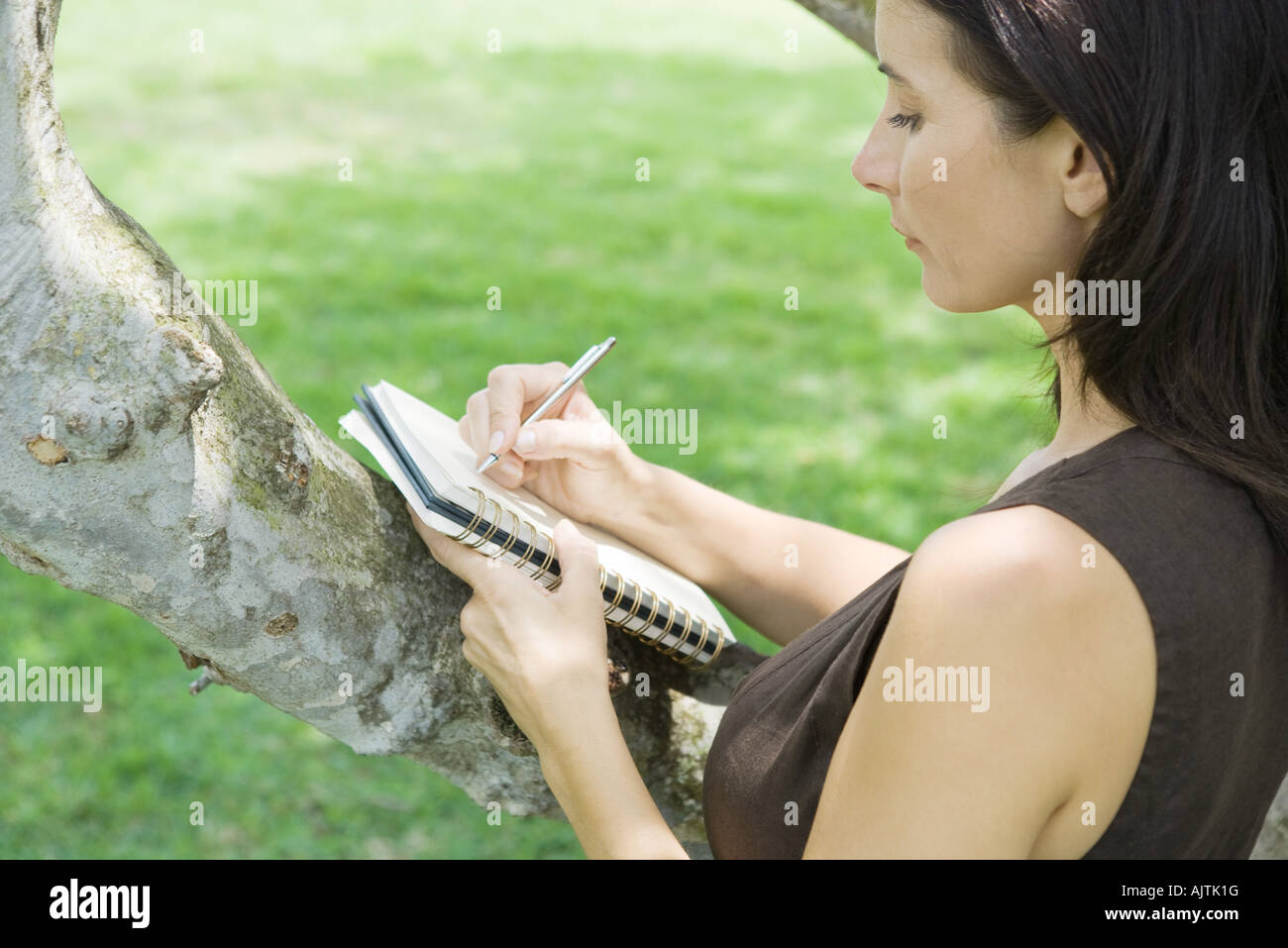 Woman writing in notebook, holding notebook against branch of tree ...