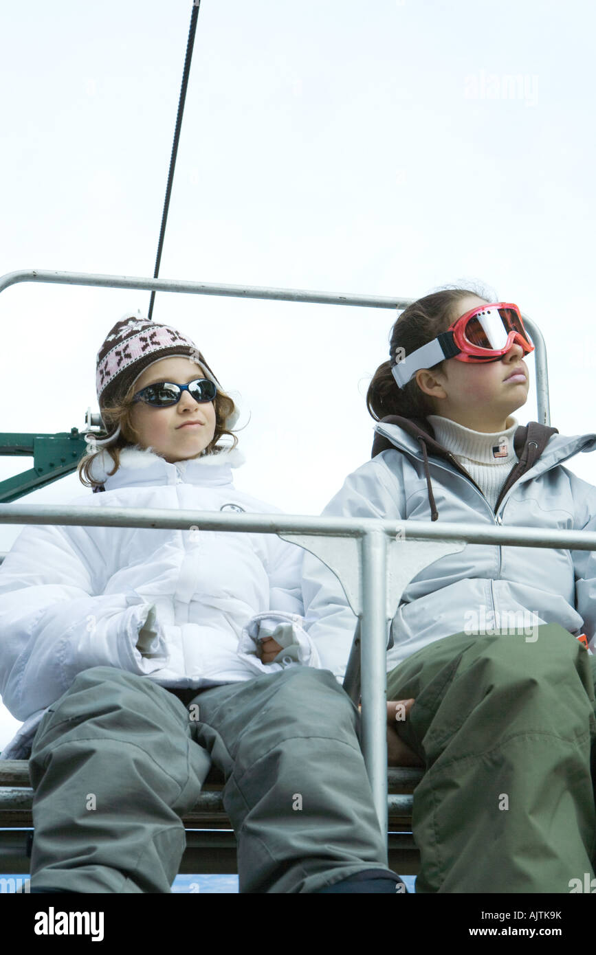 Two young friends sitting on chair lift, side by side, looking away ...