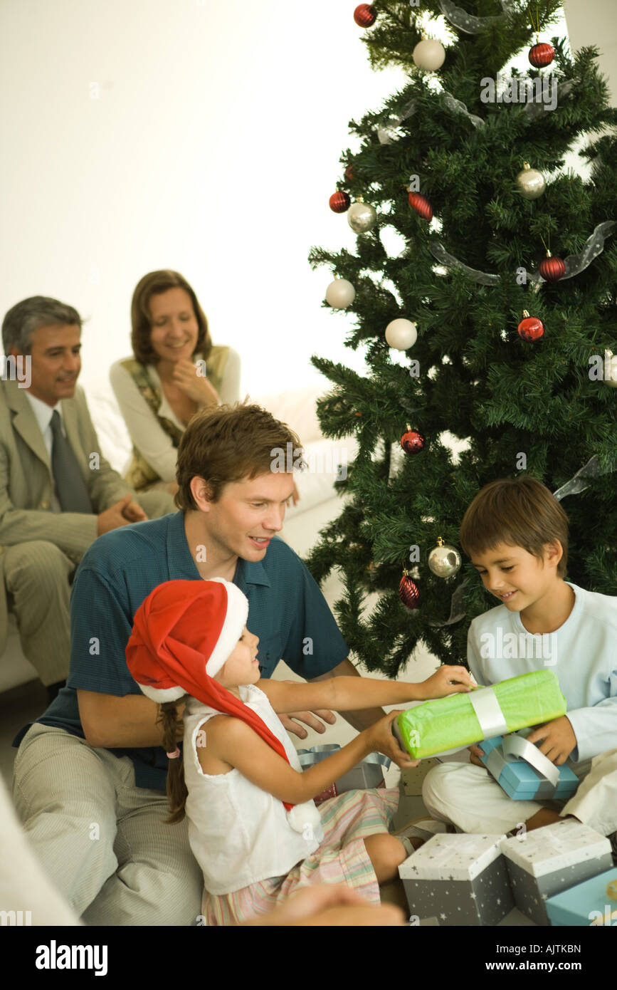 Father and two children sitting by Christmas tree, opening presents ...