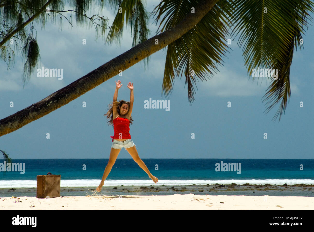 Girl stretching out jumping on white sandy tropical beach with suitcase ...