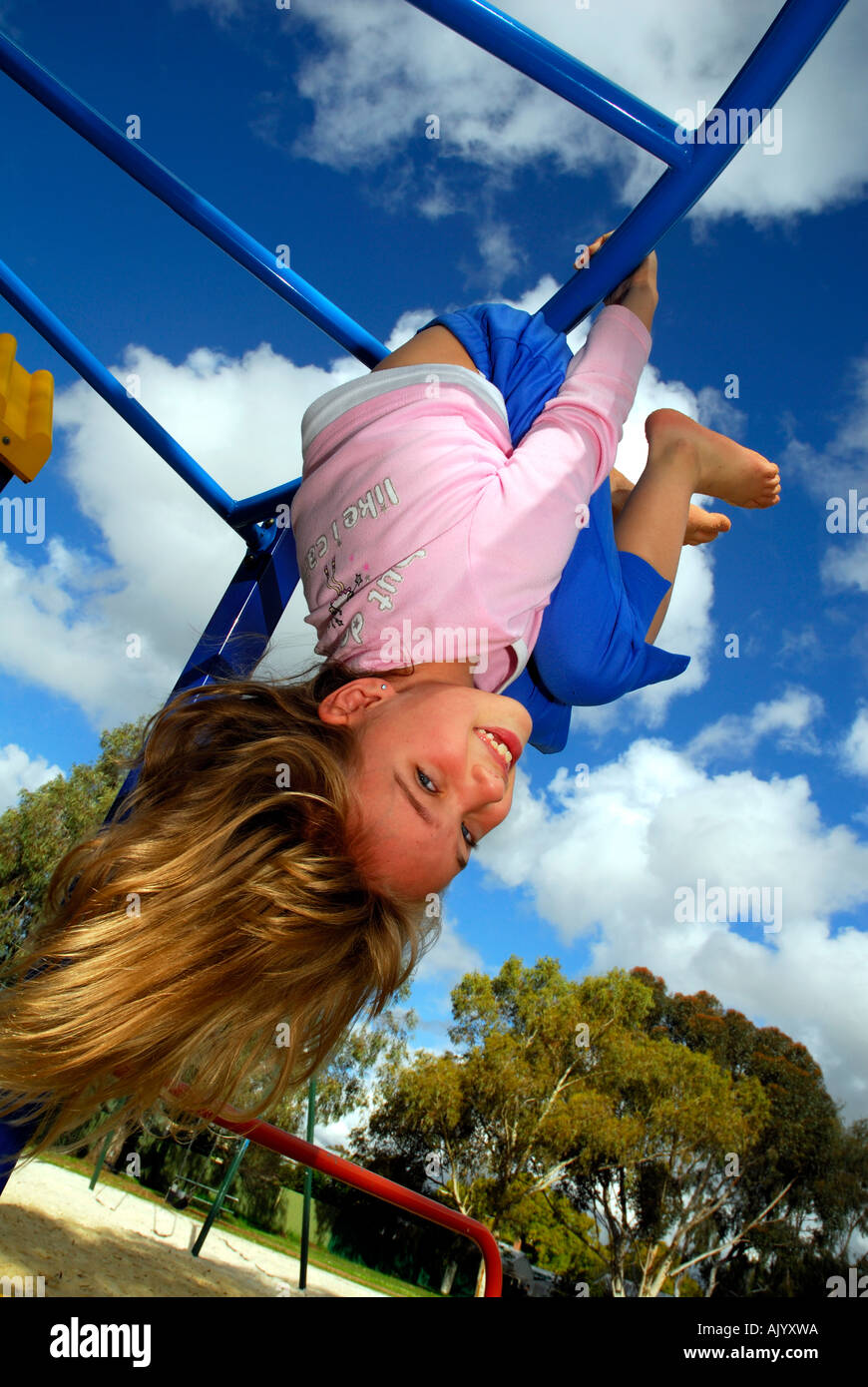 young female child hanging upside down on blue playground equipment, cloudy blue sky in ...