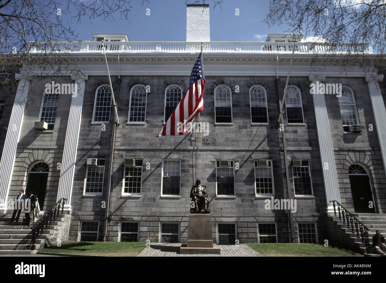 A statue of John Harvard sits under a large America flag on the campus ...