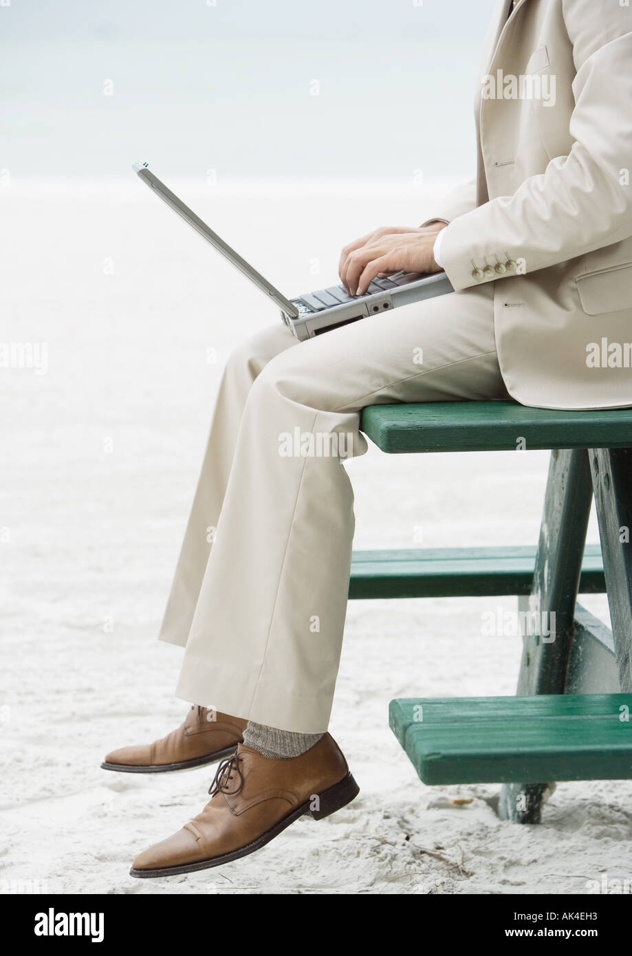 Businessman sitting on picnic table on beach, using laptop, chest down ...