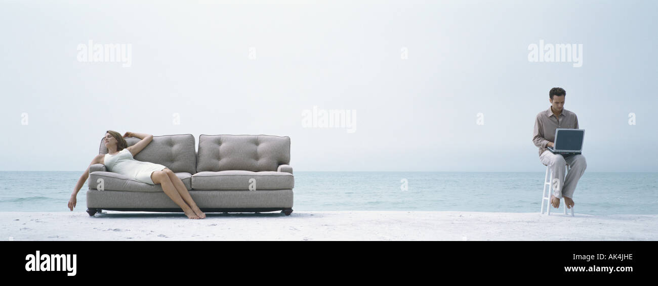 On beach, woman napping on couch while man sits on stool using computer ...