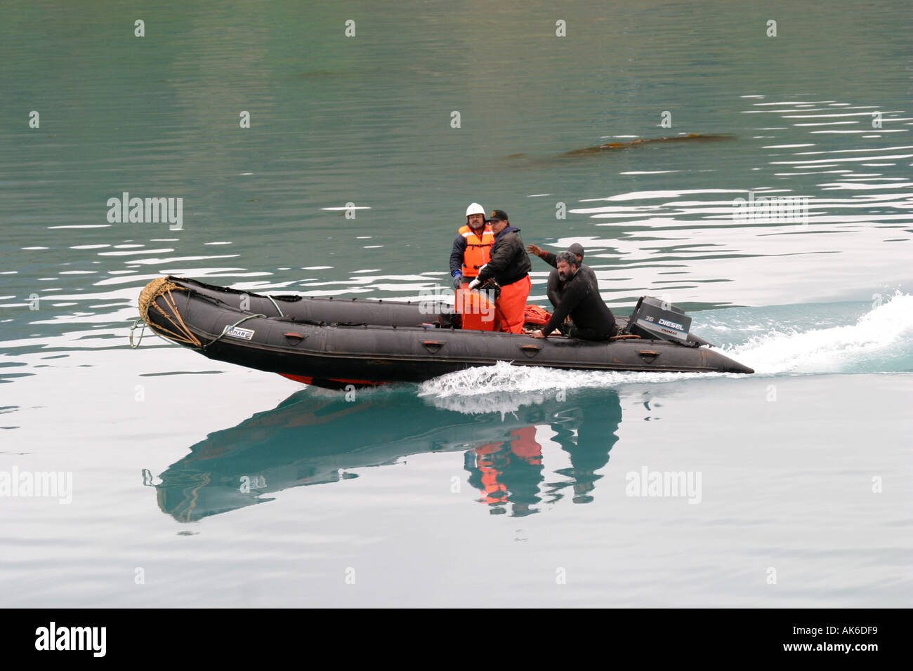 four men in a Zodiac inflatable raft reflected on still dark green ...
