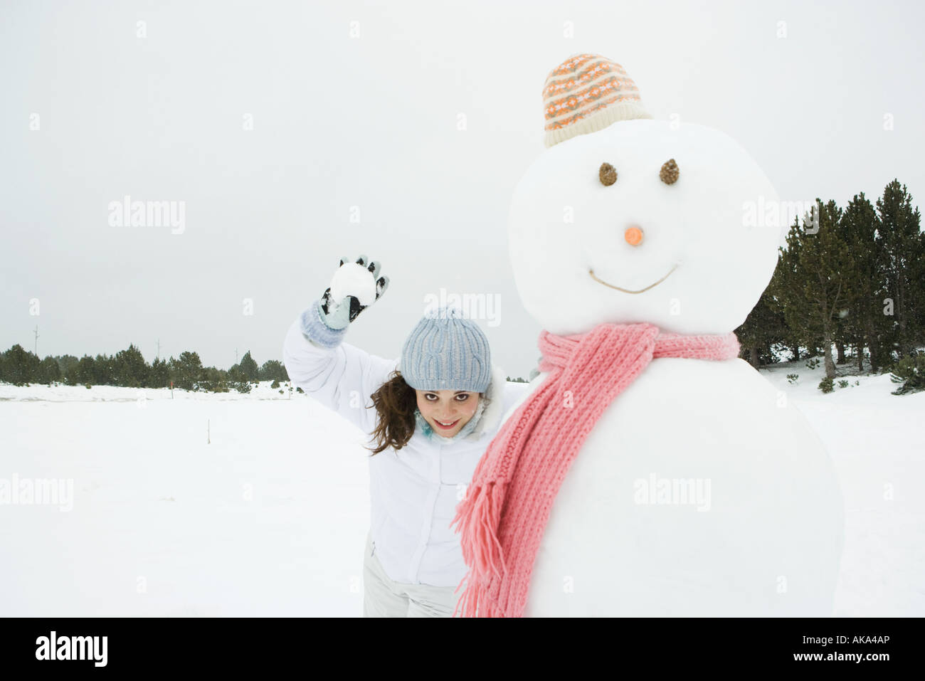 Young woman standing next to snowman, ready to throw snowball at camera ...