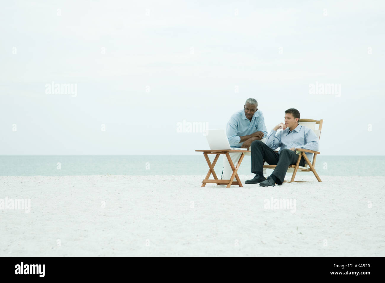 Two men on the beach, looking at laptop computer together, full length ...