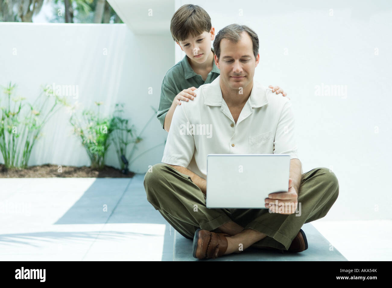 Man sitting on ground using laptop computer, son looking over his ...