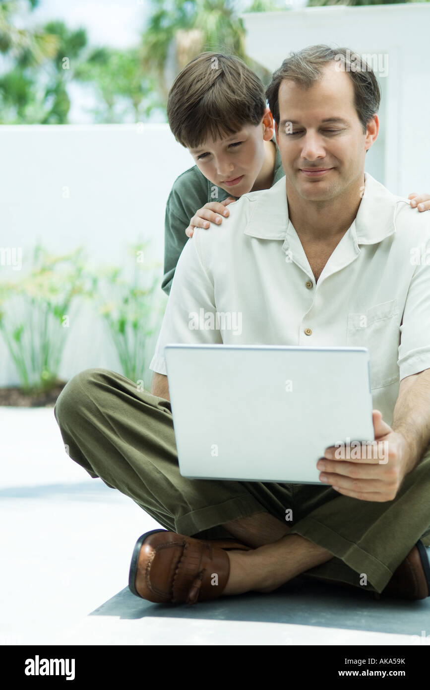 Man sitting on the ground using laptop computer, son looking over his ...