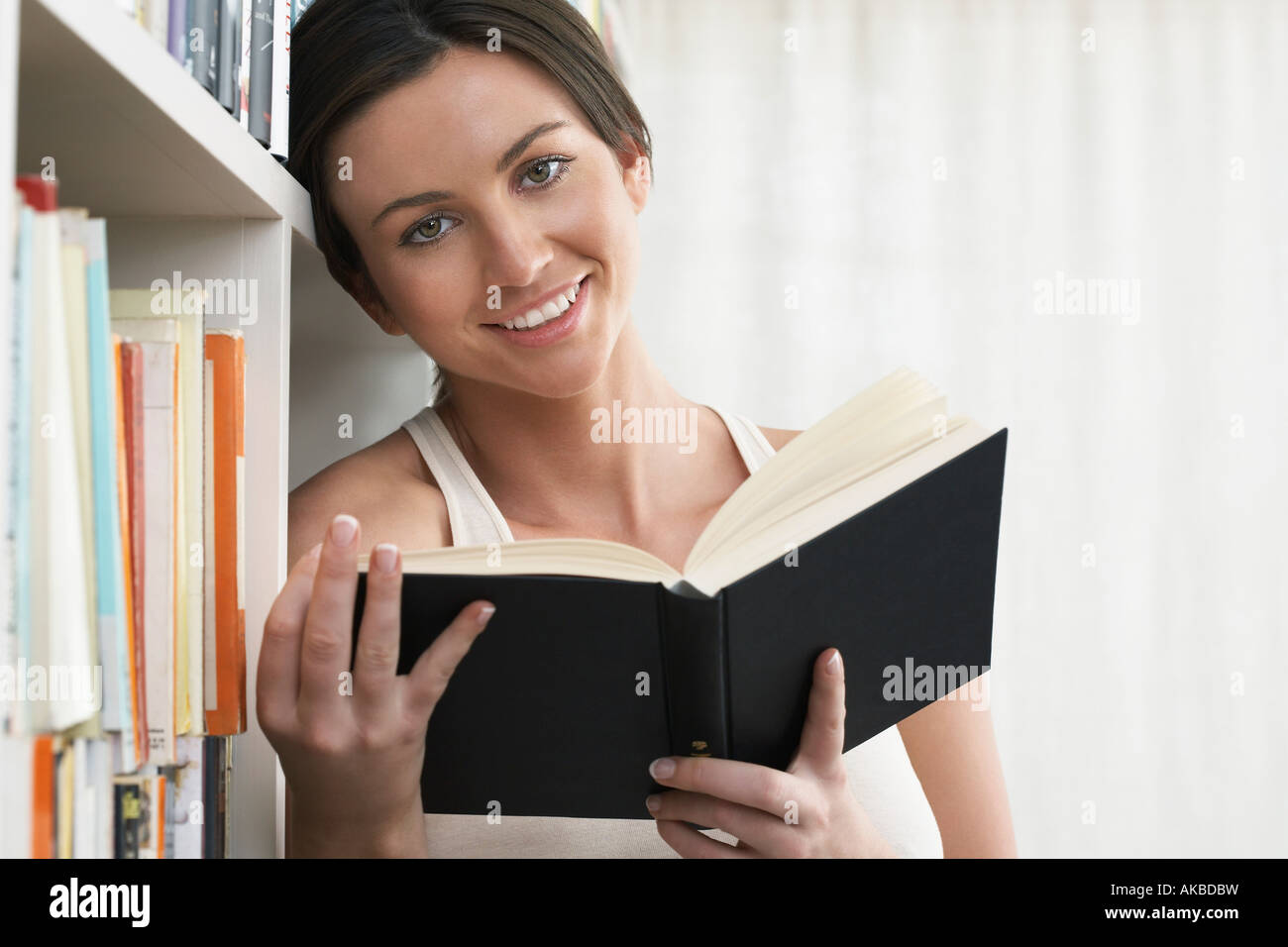 Young woman holding book, leaning against bookshelf, portrait Stock ...