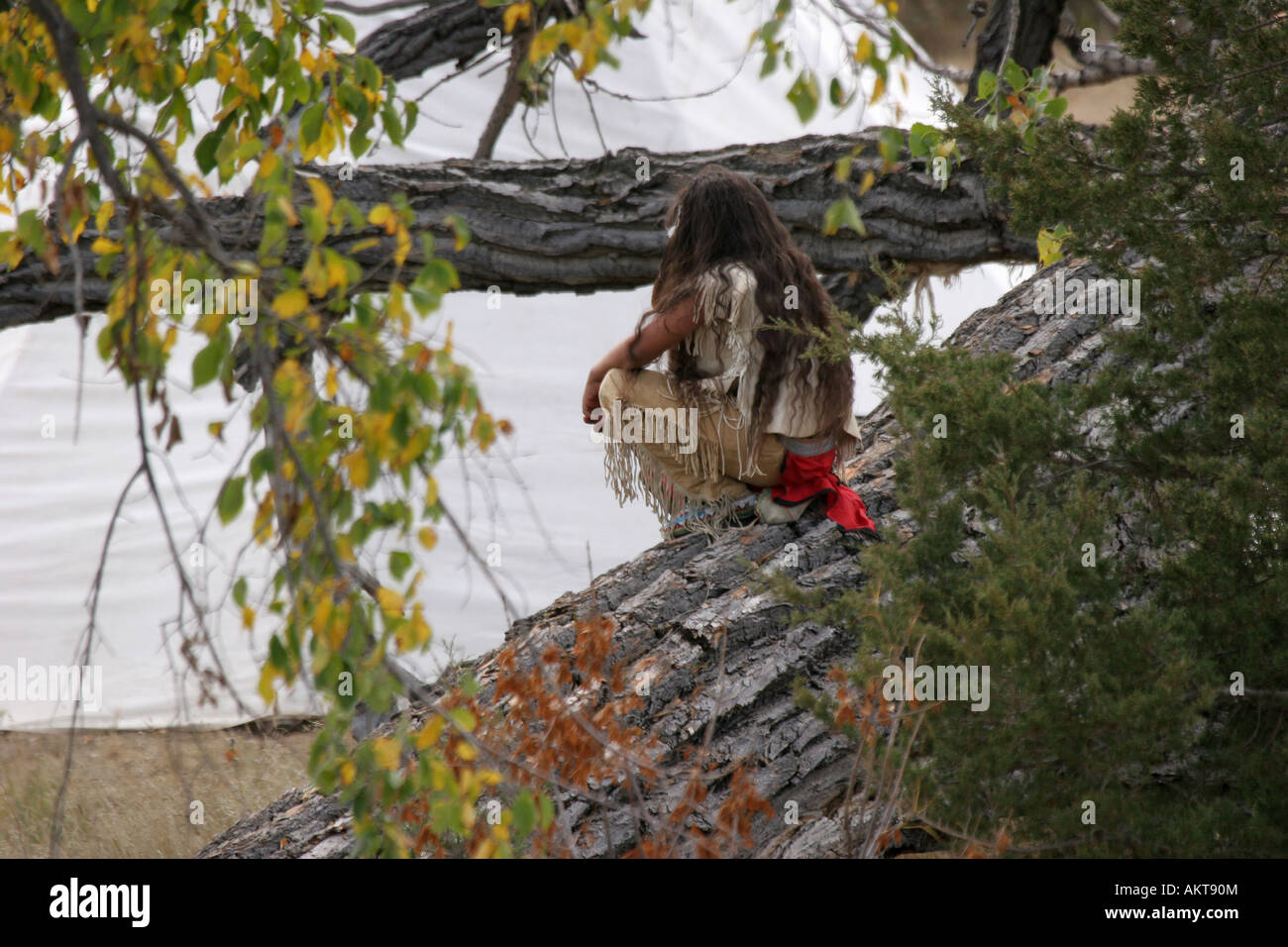 A young Native American Indian boy sitting on a downed tree in a tipi ...