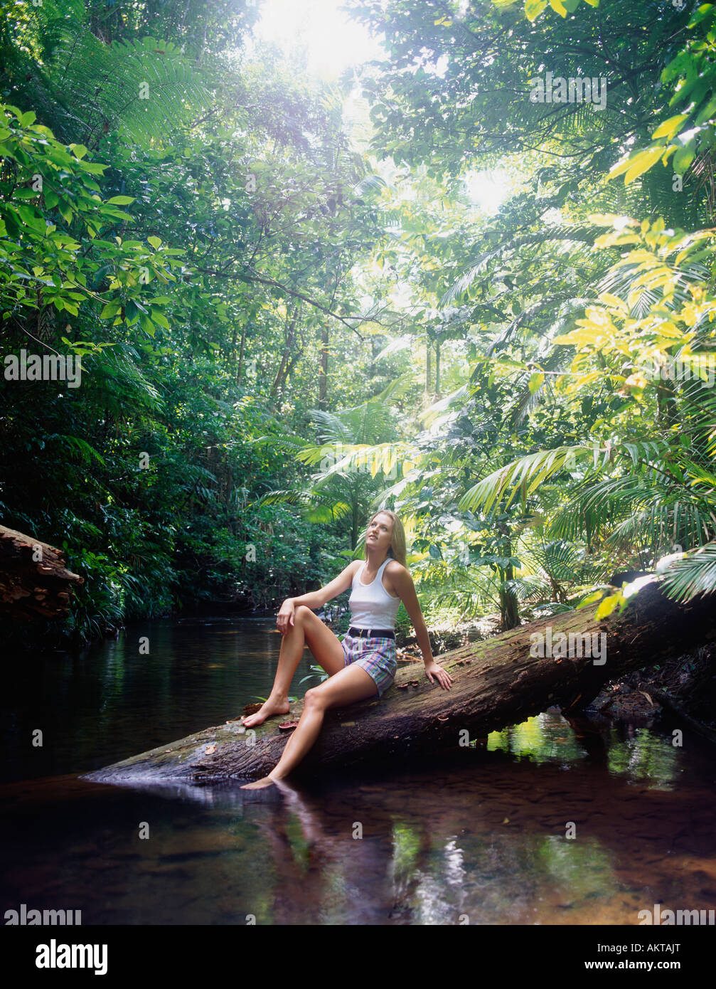 Woman sitting on a fallen tree trunk over a stream in a lush green ...